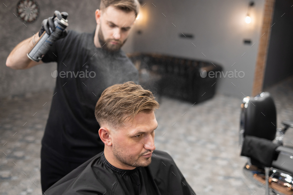 Male barber master doing hair styling to a man using hairspray Stock ...