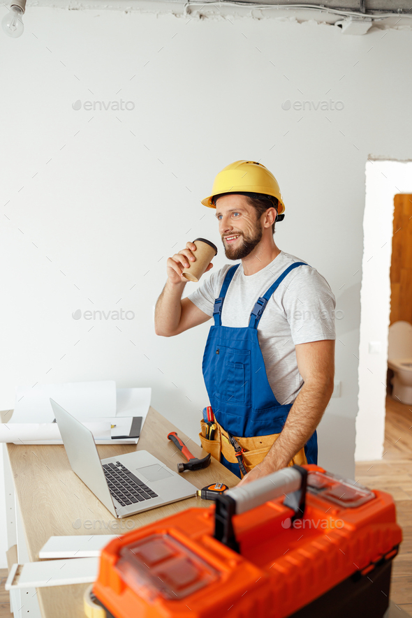 Relaxed handyman in uniform and hard hat drinking coffee during a break ...