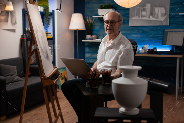 Senior artist holding laptop computer sitting in art studio with ...