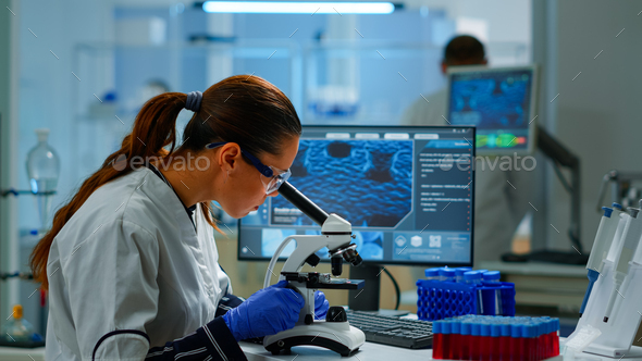 Scientist looking at biological samples under microscope typing on pc ...