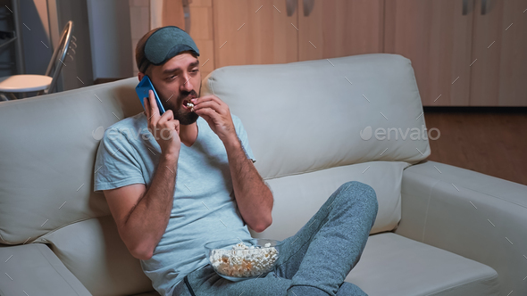 Caucasian male resting on couch eating popcorn while talking on phone ...