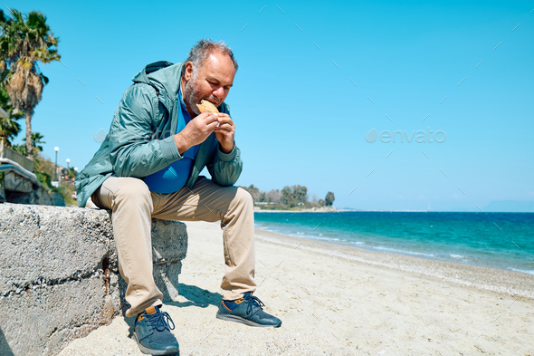 Bearded mature man at spring seaside eating hot palatable panzerotti ...