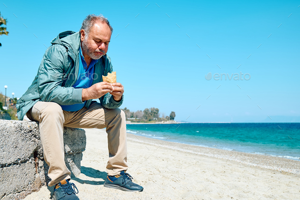 Bearded mature man at spring seaside eating hot palatable panzerotti ...