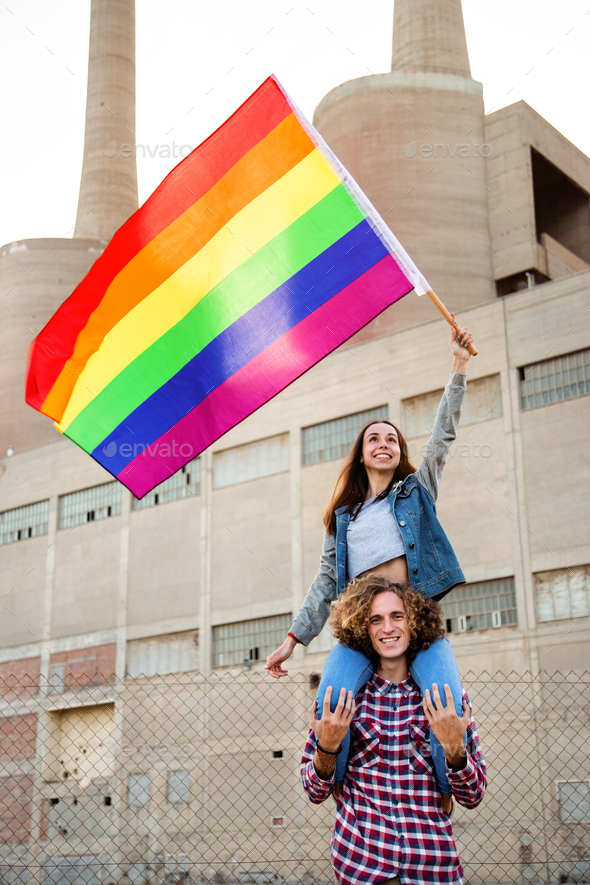 Young happy woman sitting on male friend shoulders waving rainbow flag ...