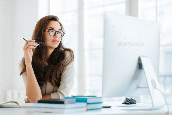 Lady focused into big monitor, sits at desktop, holds pen and writes ...