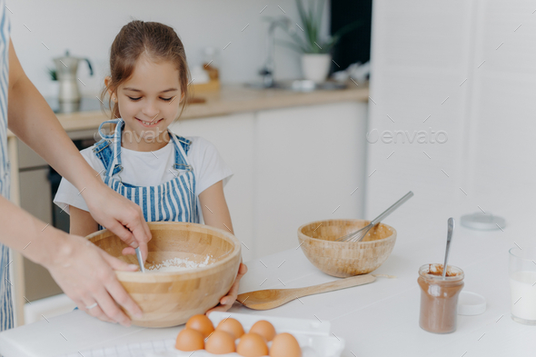 Smiling little girl helper holds big bowl, looks how mother is mixing ...
