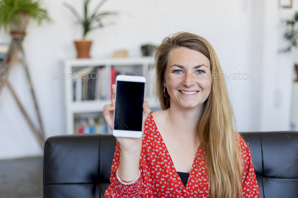 Happy lady showing a blank smart phone screen sitting on a sofa at home ...