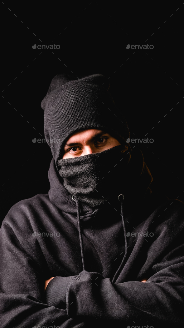 Thief in black balaclava and black cap posing with his arms crossed ...