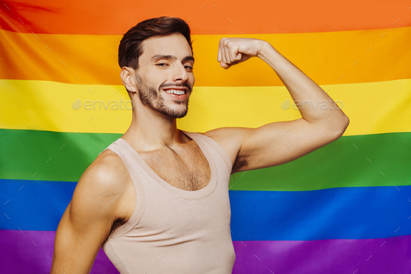 Cheerful gay man showing biceps muscle over rainbow flag background ...