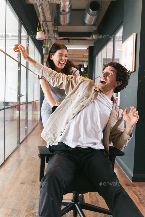 Young woman having fun pushing man on chair along walkway in open plan ...
