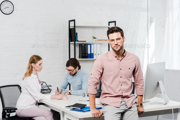 handsome young businessman looking at camera and leaning back on table ...