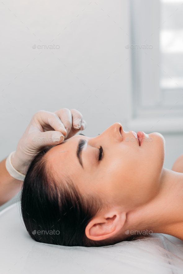 partial view of cosmetologist putting needle on womans forehead during ...