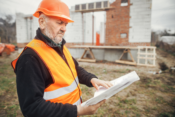 Senior male foreman in hardhat checking blueprints of building new ...