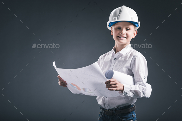 portrait of smiling boy in architect helmet holding blueprints on dark ...