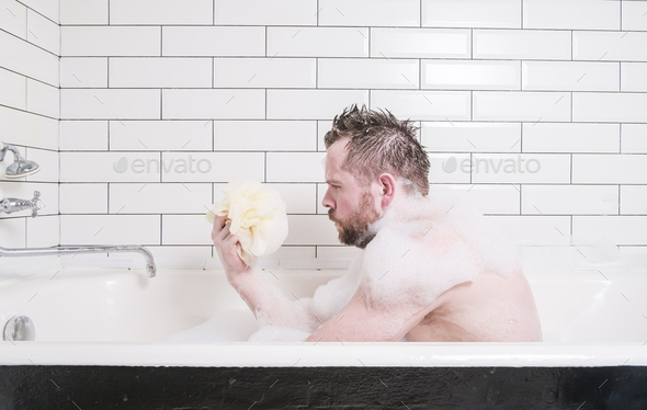 Sad man bathes in a bath with lush soap suds, he holds a sponge in his ...