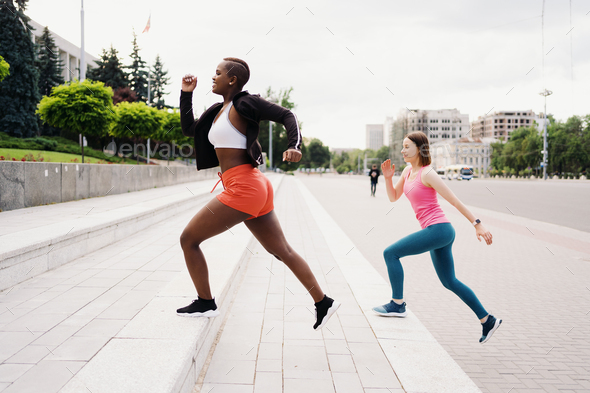 Two multiethnic girls running through the city streets, outdoor ...