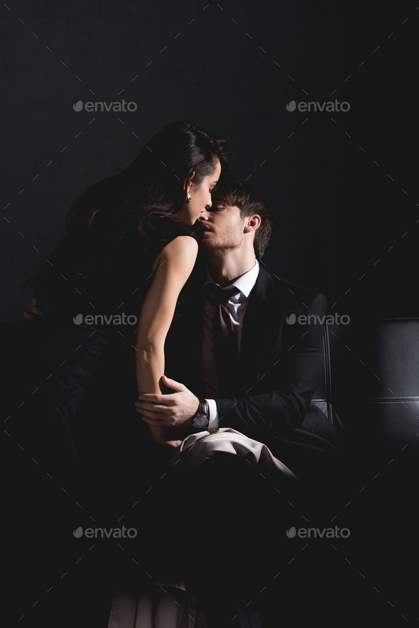 man in suit and woman in black dress kissing on couch on dark ...