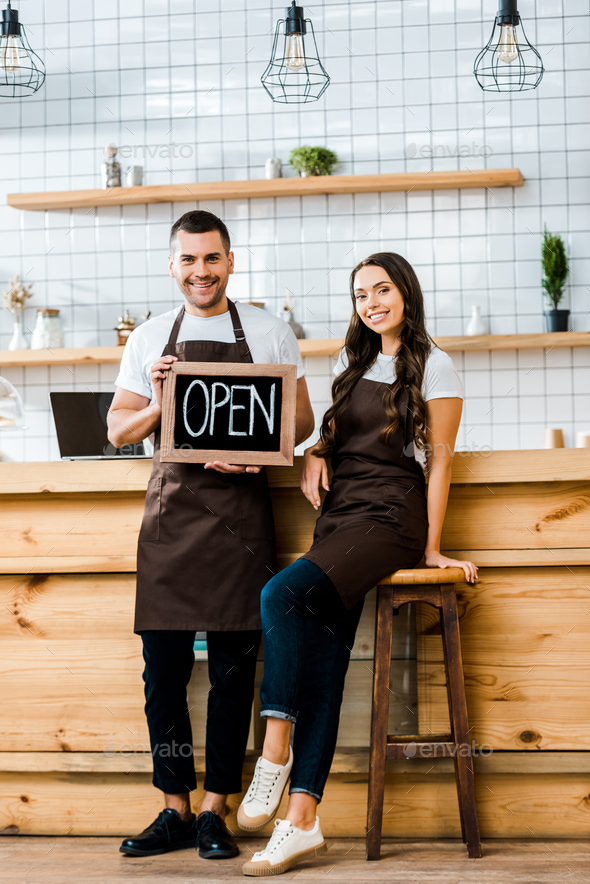 cashiers in aprons standing near wooden bar counter and holding ...