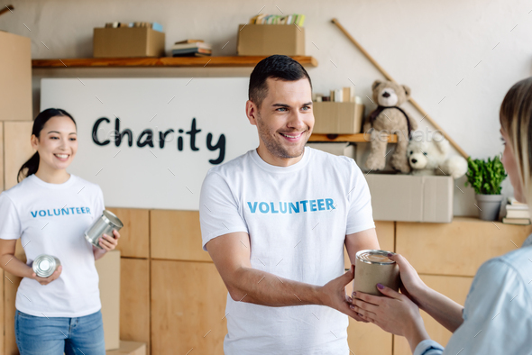 young, smiling multicultural volunteers giving canned food to woman in ...