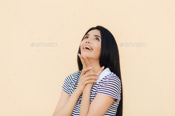 Female model looks happily upwards and keeps hand under chin, notices ...