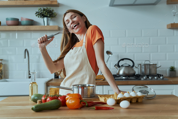 Playful young woman using whisk like microphone while cooking at the ...