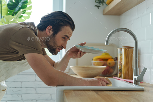 Handsome young man checking dishes after washing while standing near ...