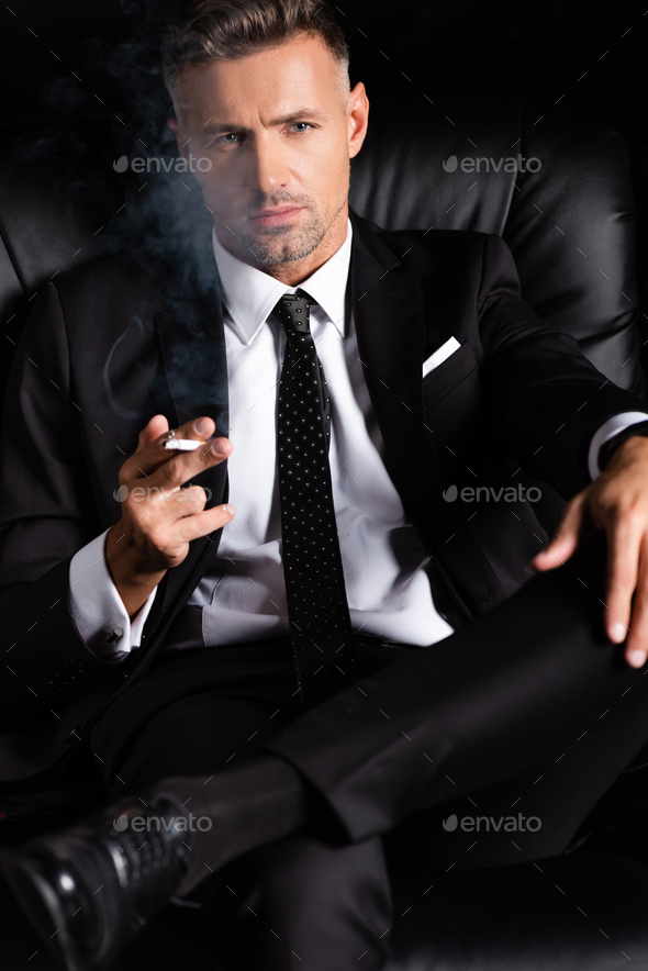 Confident businessman looking at camera and smoking cigarette on sofa ...
