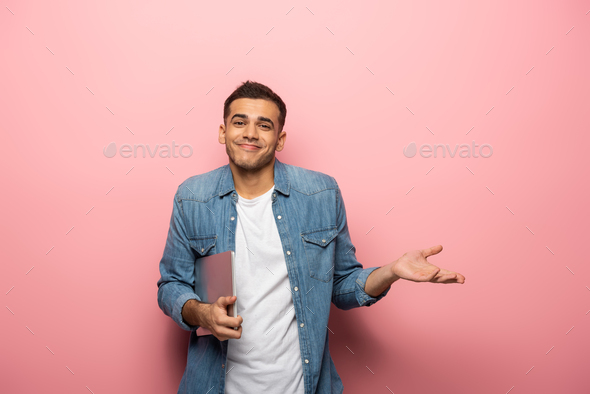 Young smiling man with laptop showing shrug gesture on pink background ...