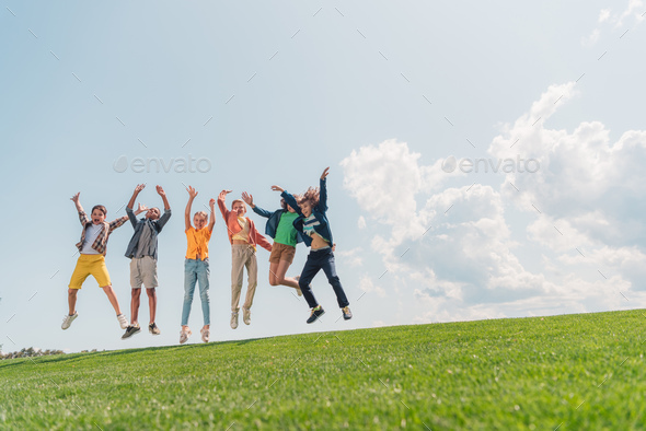 cheerful multicultural kids jumping and gesturing against blue sky ...