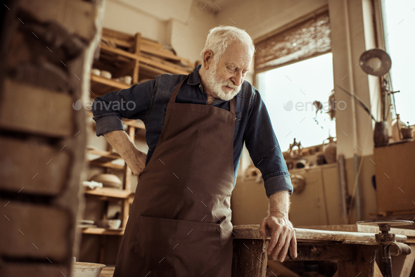 Front view of senior potter in apron standing and leaning on table ...