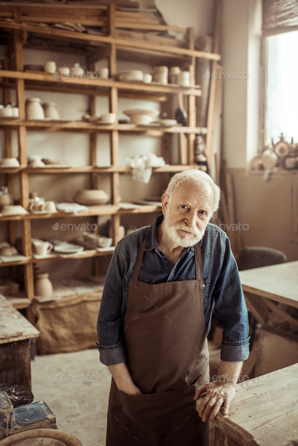 Front view of senior potter in apron standing and leaning on table ...