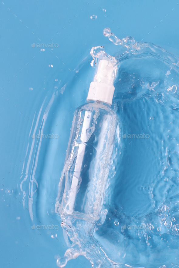 Plastic spray bottle of cosmetic product and splashing water on blue ...