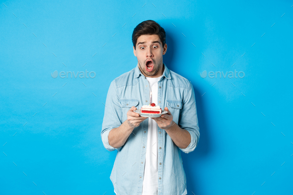 Man looking shocked at birthday cake, standing against blue background ...