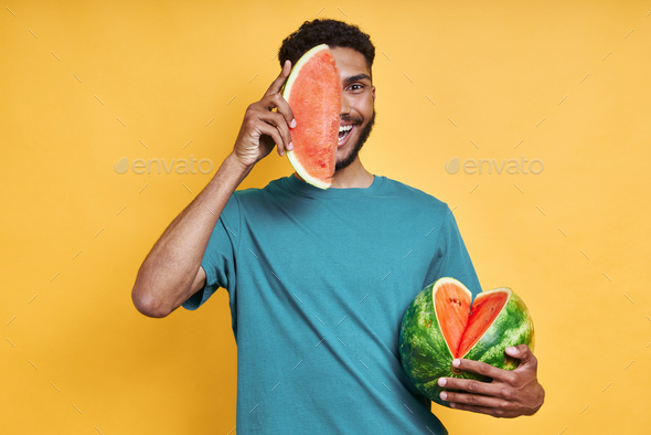 Playful African man holding slice of watermelon near face while ...