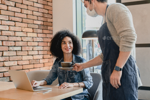 Portrait of a waiter serving coffee to african customer with laptop in ...