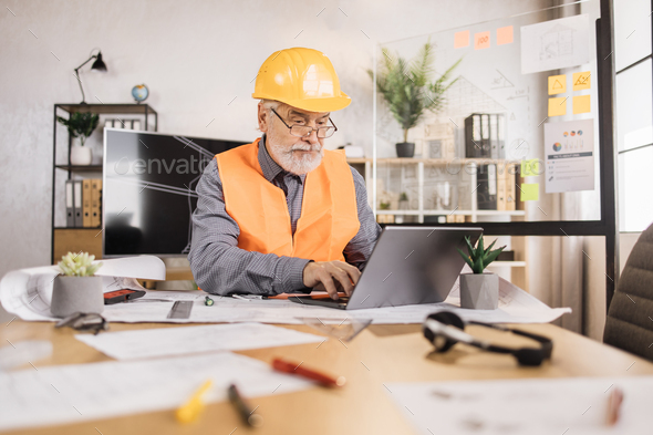 Engineer senior man in helmet and reflective vest sitting at table with ...