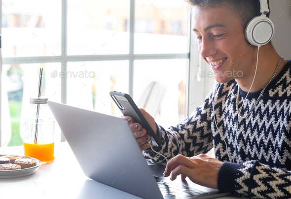 Smiling teenage boy using laptop computer listening to music from smart ...