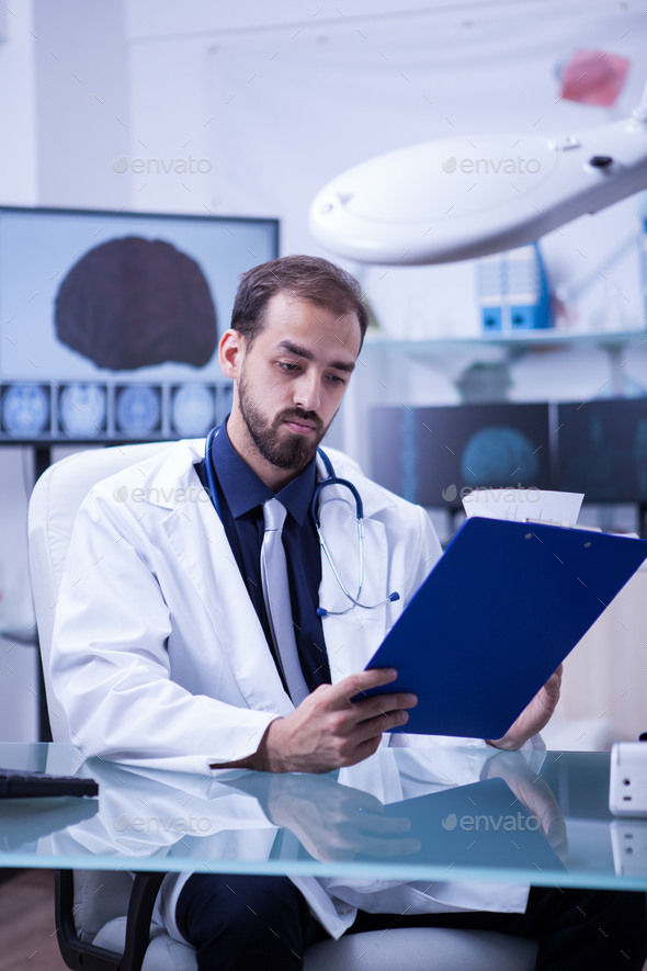 Specialist doctor checking his clipboard sitting at his desk in the ...