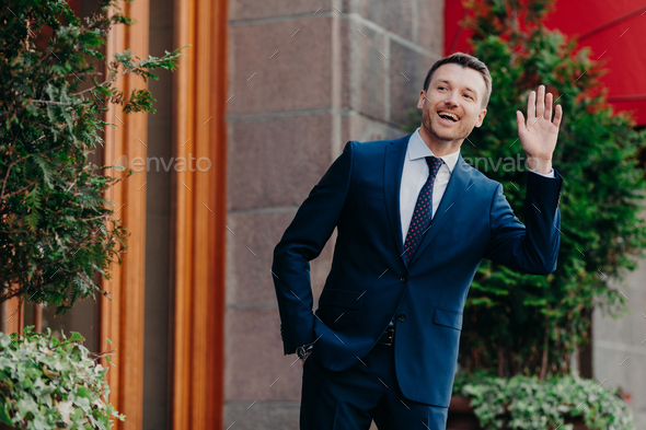 Shot of cheerful male banker in luxury black suit, waves with hand as ...