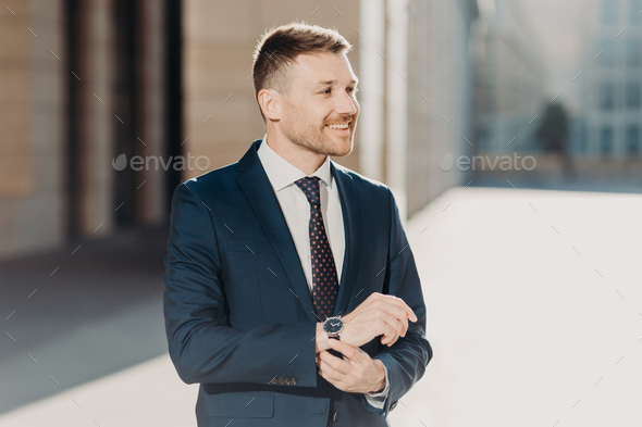 Sideways shot of happy male manager in formal suit, keeps hand on watch ...