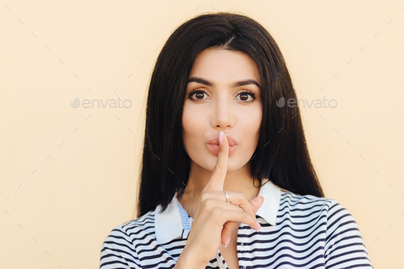 female with silence sign keeps fore finger on lips, asks to be quiet as ...