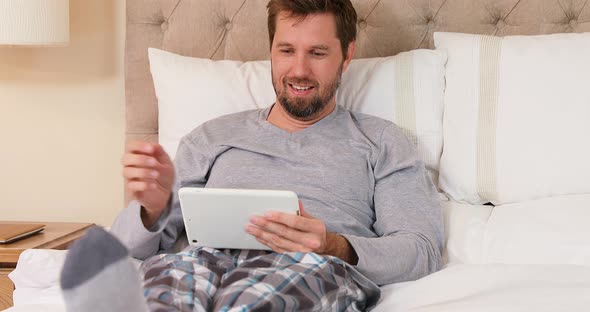 Smiling young man sitting on bed using his tablet in the bedroom 4K 4k ...