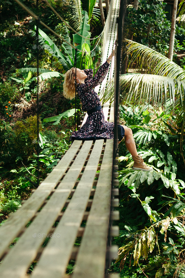 side view of attractive woman posing on suspension bridge in jungle ...