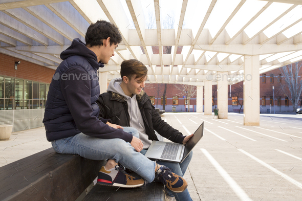 side view couple of student friends sitting on a bench looking at a ...