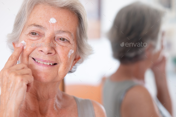 Portrait of senior beautiful gray-haired woman applies anti aging cream ...
