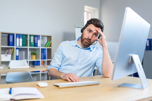 Tired man working at a computer with a headset for a video call in the ...