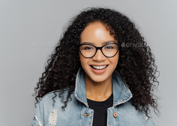 Glad woman with curly hair, wears optical glasses, denim jacket, looks ...