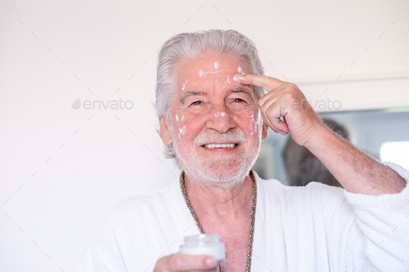 Skin care. Handsome elderly bearded man in bathrobe applying cream on face as daily routine ...