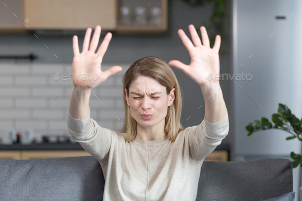 Scared woman sitting on sofa at home, holding hands in front, stop sign ...
