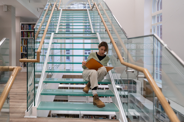 Curious female reader sits on modern glass stairs step reading favorite ...